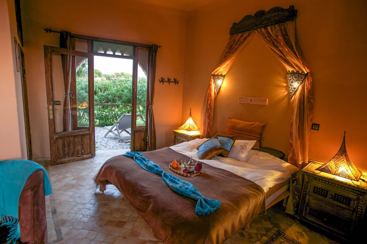 Quarto com cama, mesa de cabeceira, abajur e vista para o exterior.