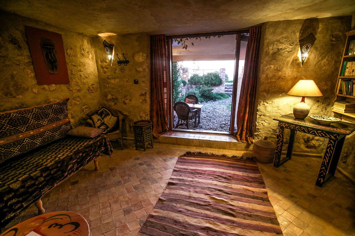 Quarto aconchegante com sofá, mesa pequena com abajur, tapete estampado e vista para o exterior pela janela.
