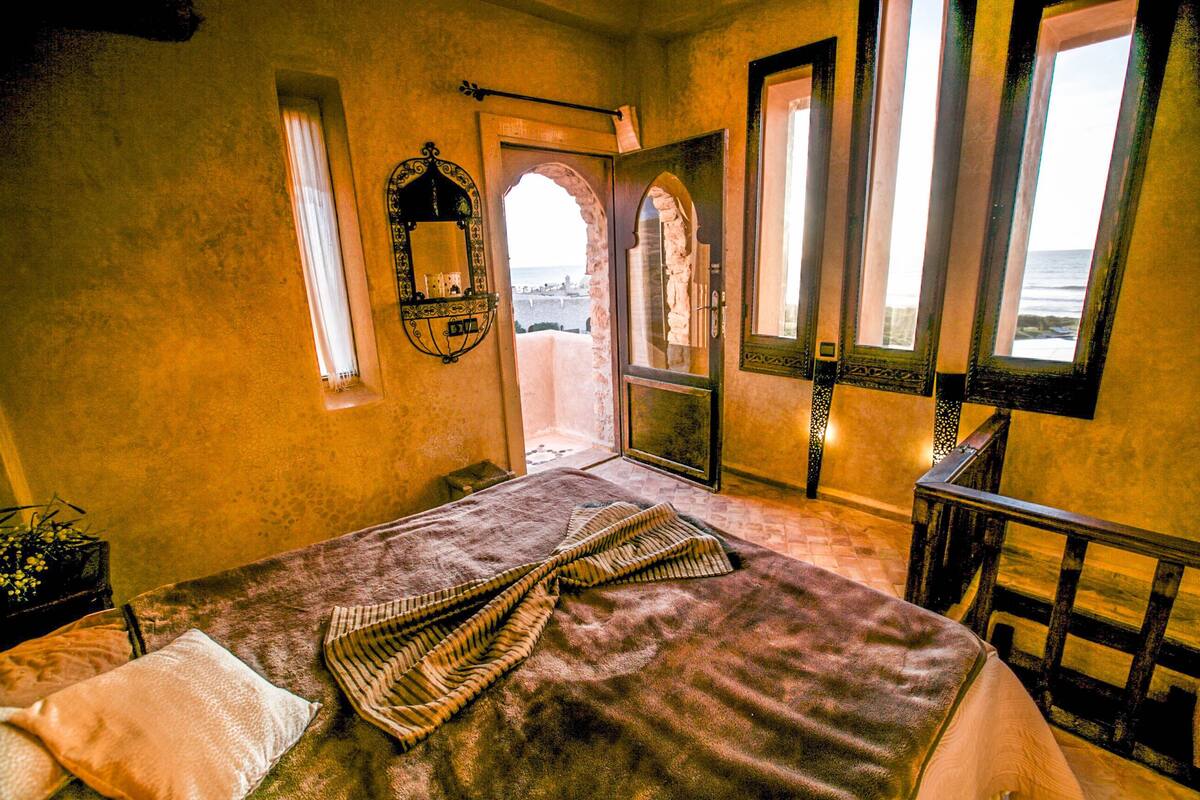 Quarto com cama, janela com vista, porta e uma pequena mesa com uma planta.