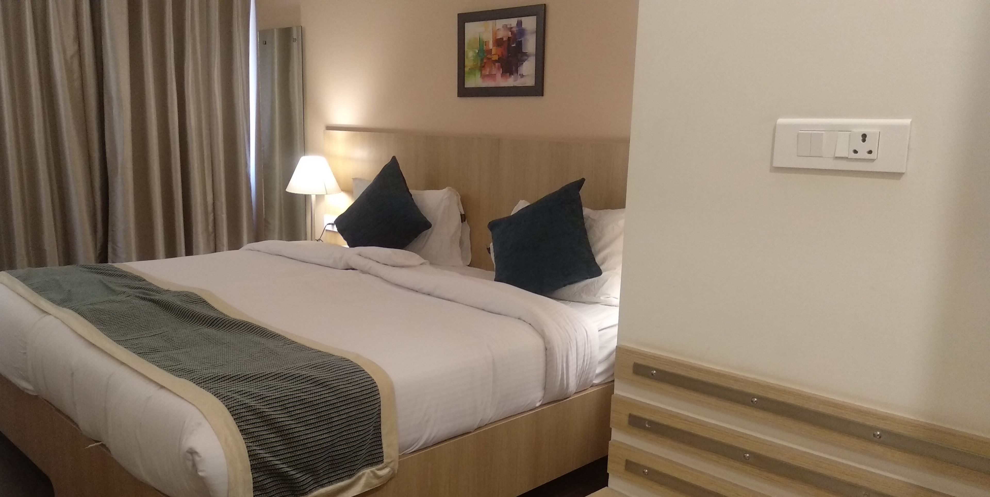 Quarto de hotel com uma cama, uma luminária, um quadro na parede e uma tomada.