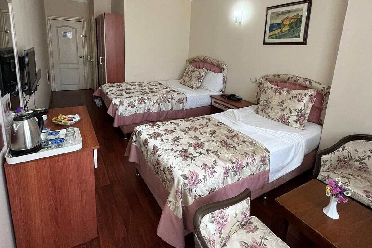 Quarto de hotel com duas camas, uma secretária com chaleira, uma cadeira e uma pequena mesa com um vaso.