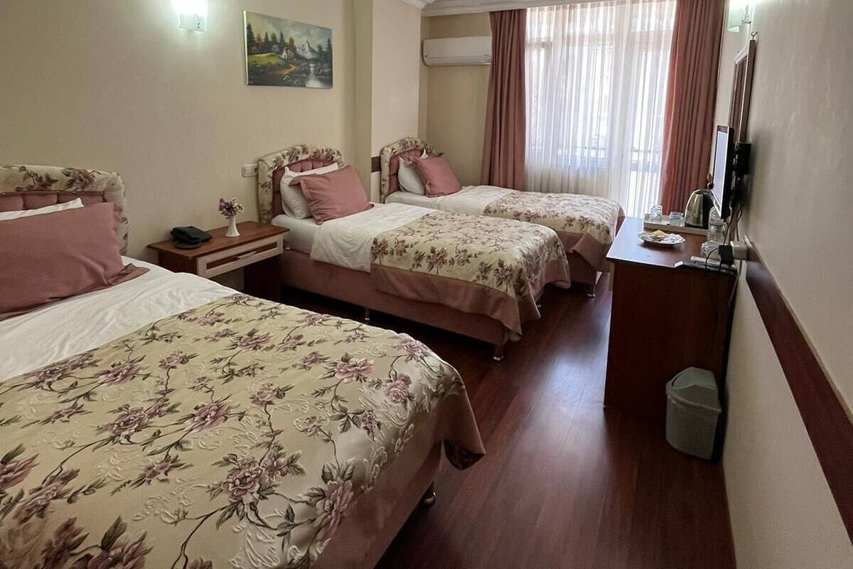 Quarto de hotel com três camas, uma escrivaninha, um cesto de lixo e um quadro na parede.