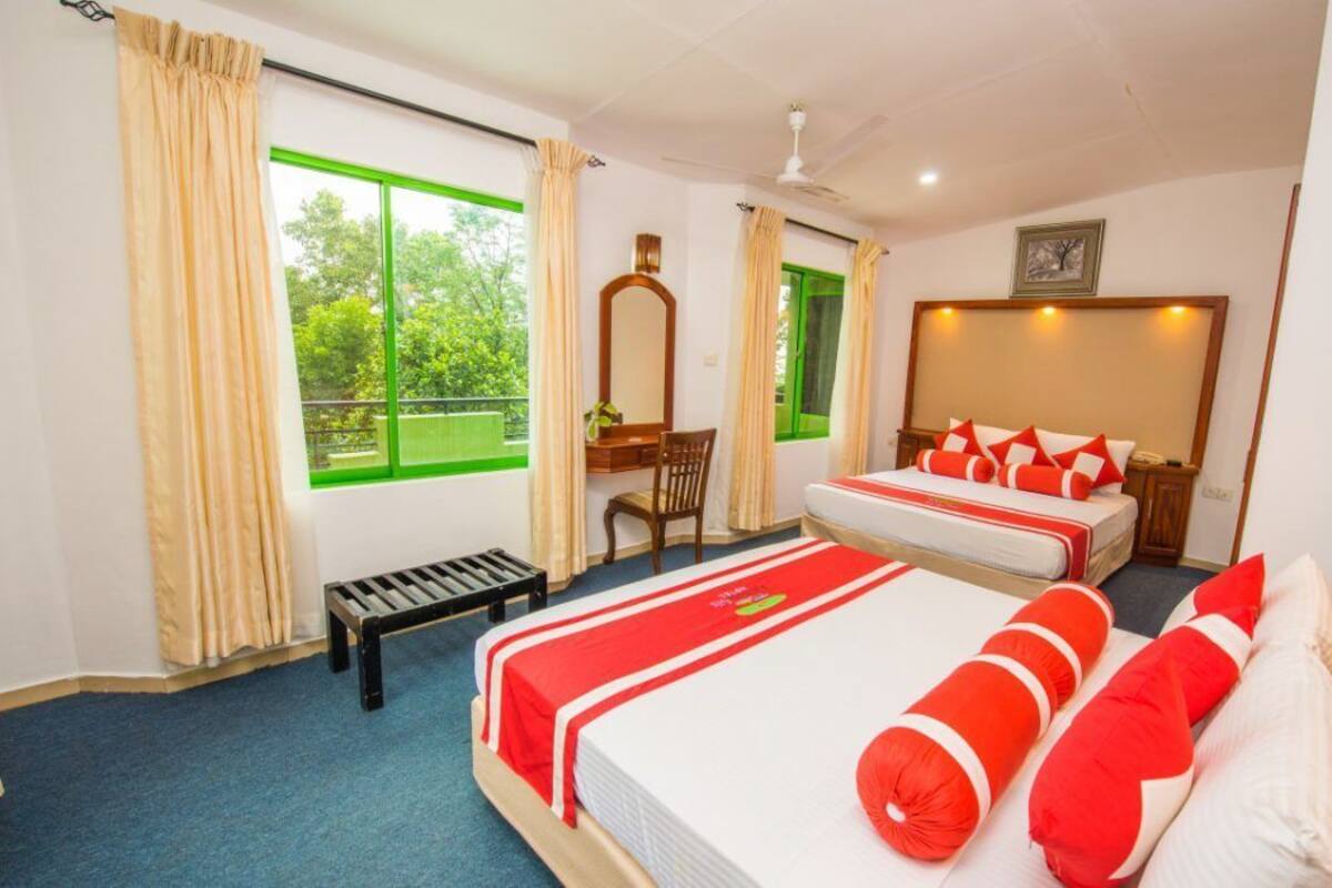 Quarto de hotel com duas camas, uma escrivaninha, uma cadeira, um banco e amplas janelas com cortinas.