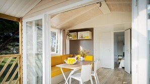 Uma sala de estar moderna e espaçosa, com um deck de madeira, um sofá amarelo, uma mesa de jantar com cadeiras, e uma cozinha visível por uma porta aberta.