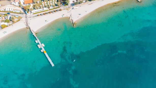 Vista aérea de uma praia com um píer que se estende sobre a água, repleto de barcos e uma fileira de edifícios à beira-mar.