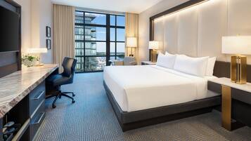 Quarto de hotel com uma cama grande, uma escrivaninha com cadeira, televisão e vista para a paisagem urbana através de janelas amplas.