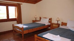 Quarto com duas camas de madeira, cada uma com uma mesa de cabeceira e uma luminária. Há uma janela com cortinas no lado esquerdo.