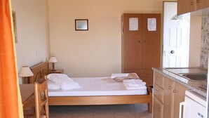 Quarto pequeno e arrumado com uma cama de solteiro, um guarda-roupa de madeira, uma mesa pequena com uma cadeira e uma área de cozinha compacta.