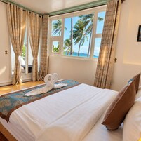 Quarto de hotel com cama, cortinas, janela vista para palmeiras e mar, e toalha sobre a cama.
