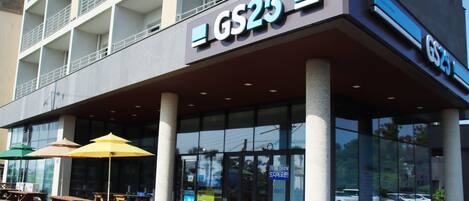 Um edifício com a placa GS25, com área externa de estar com guarda-sóis, grandes janelas e estacionamento visível.
