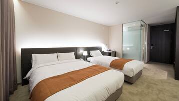 Quarto de hotel com duas camas, uma escrivaninha, uma cadeira, uma mesinha de cabeceira e uma porta de vidro que dá para o banheiro.