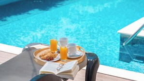 Cenário junto à piscina com uma bandeja de croissants, suco de laranja e um livro.