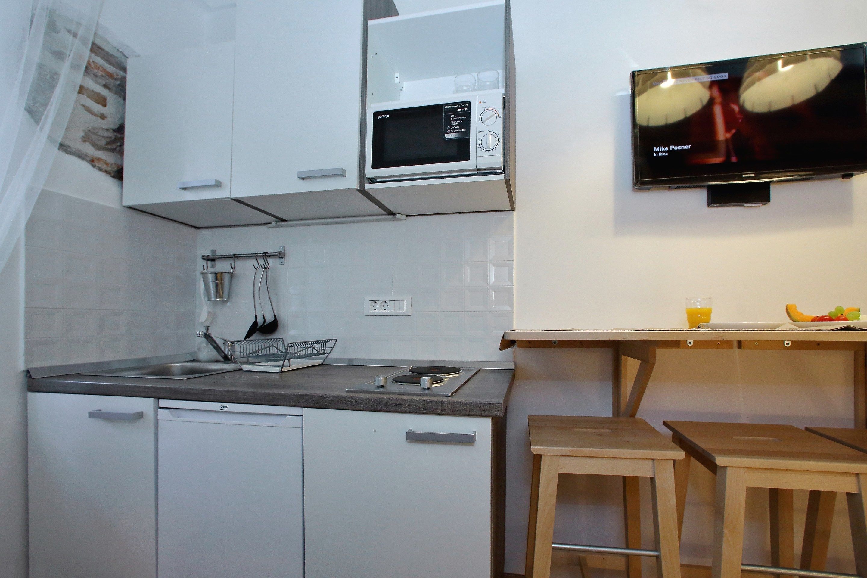 Cozinha compacta com armários brancos, micro-ondas, fogão e televisão fixada na parede.