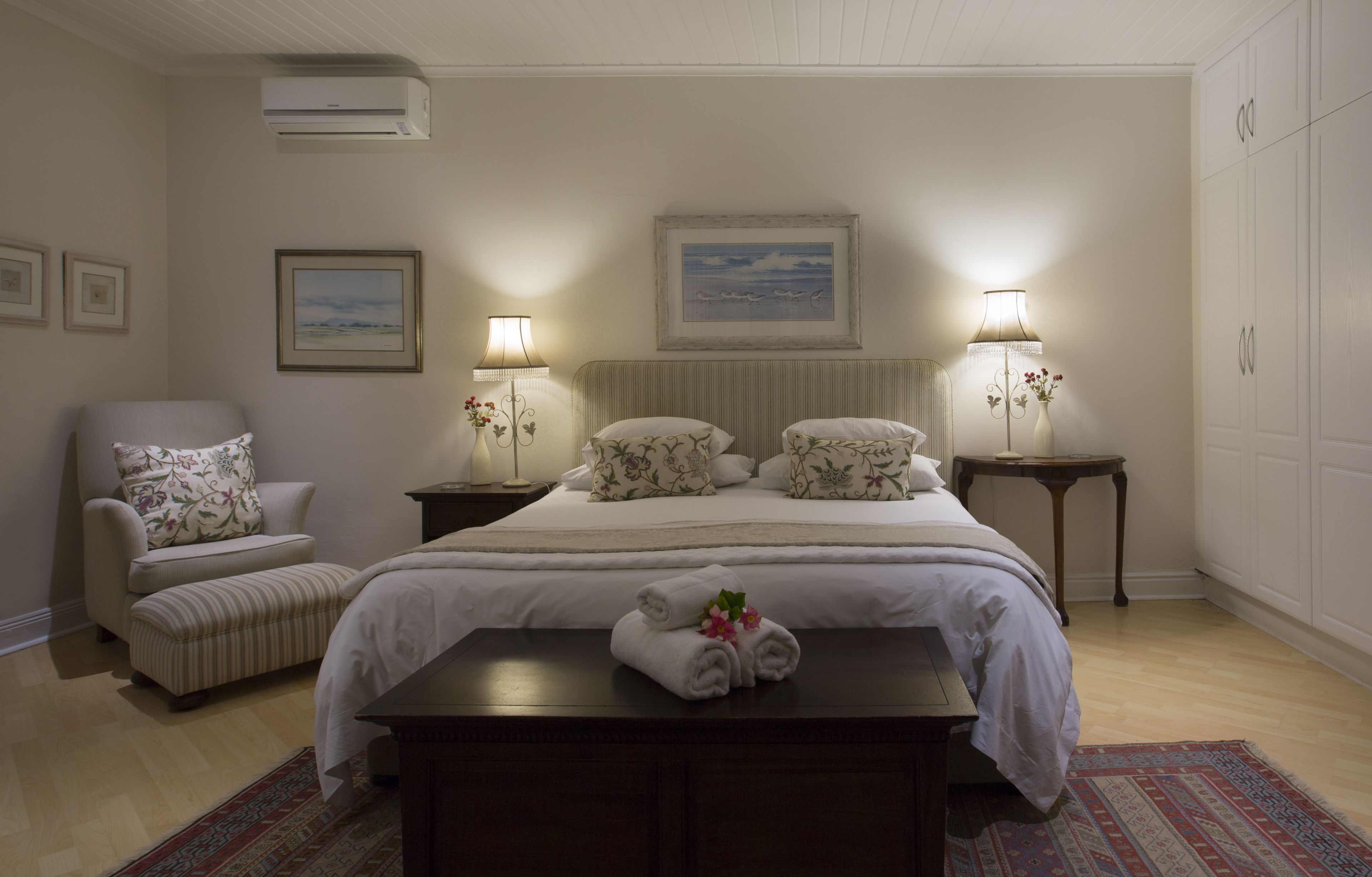 Um quarto bem arrumado com uma cama, duas mesas de cabeceira com luminárias, uma cadeira e uma pequena mesa com toalhas.