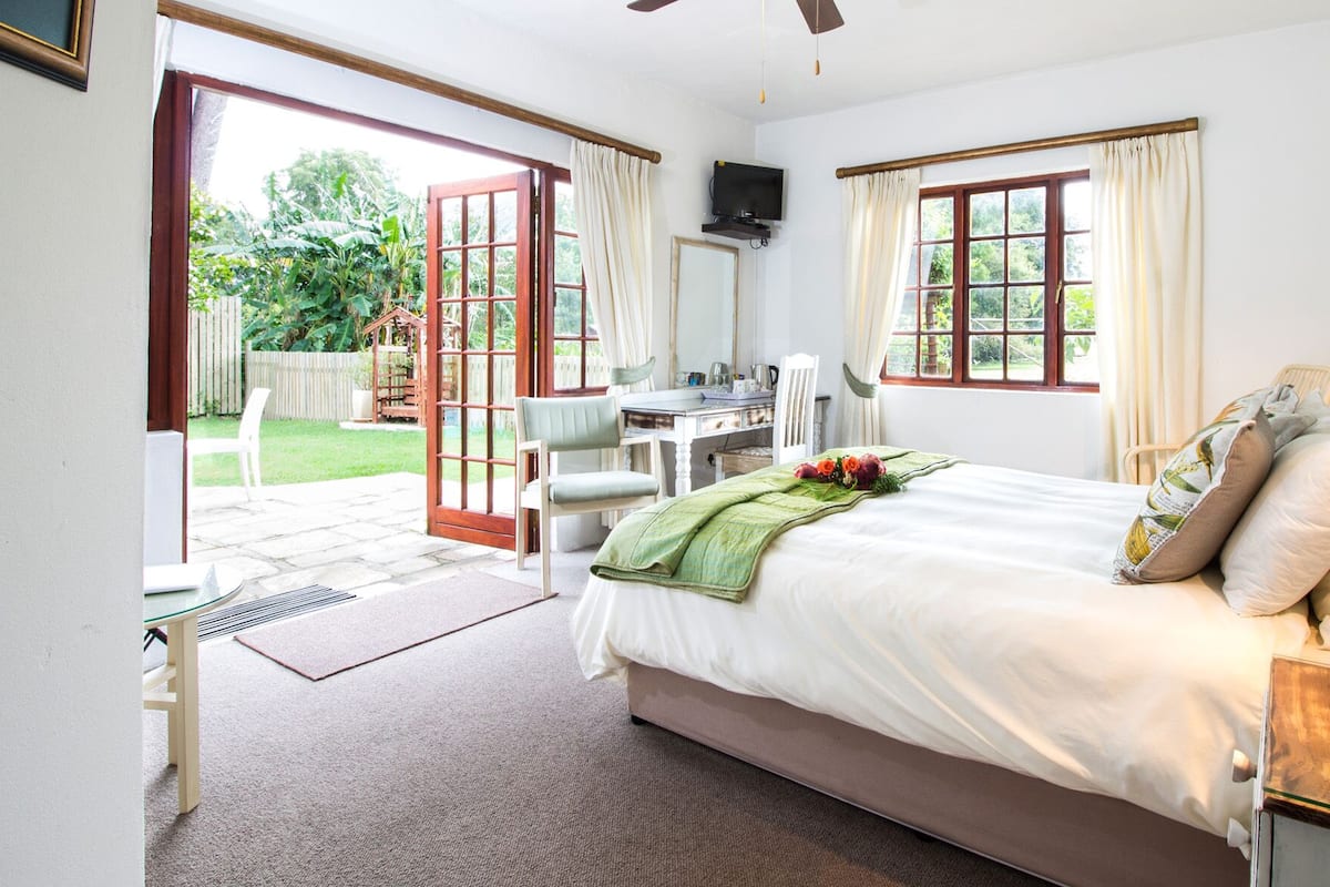 Quarto com cama, escrivaninha e cadeira. Há uma janela com cortinas e vista para o jardim.