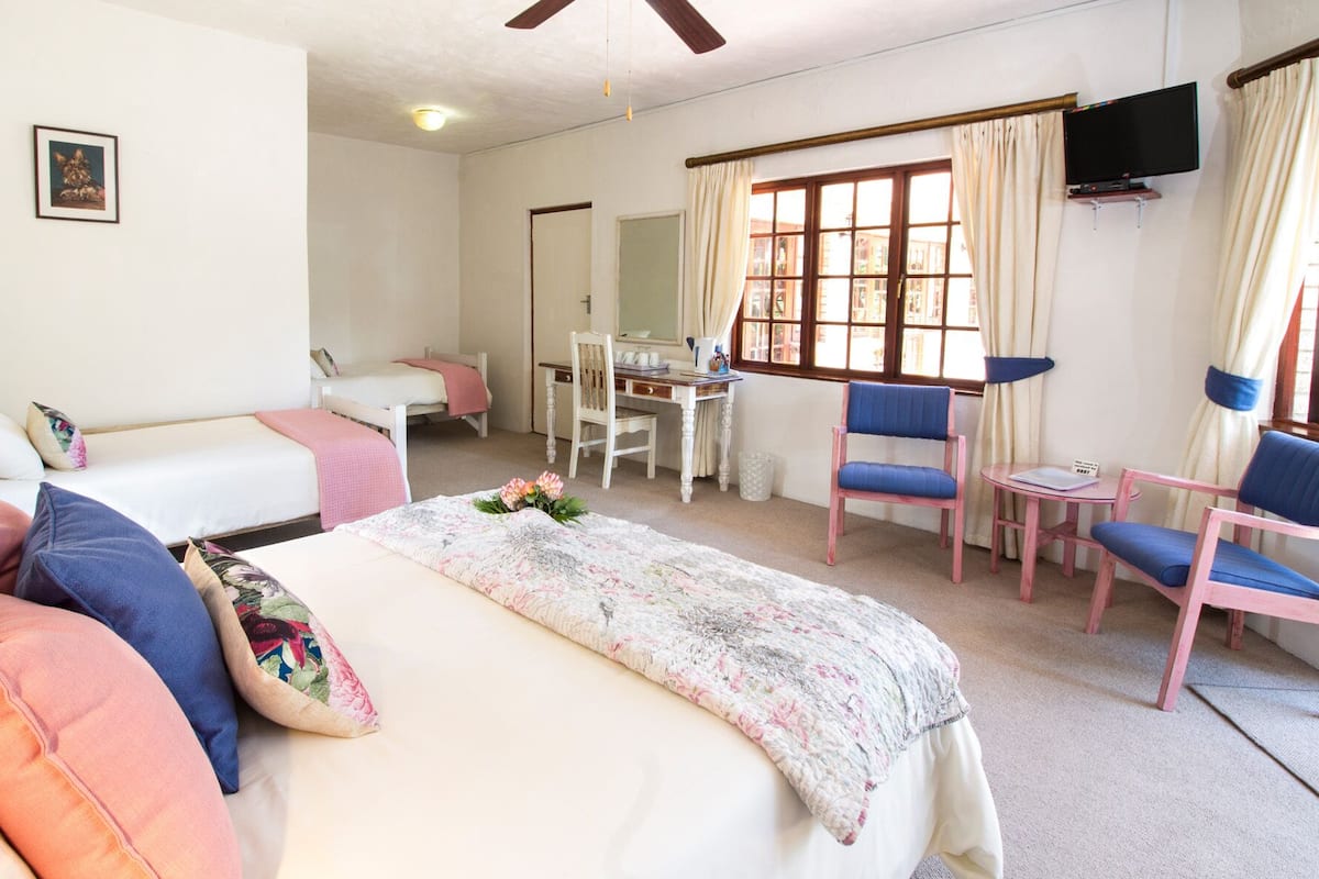Quarto de hotel com cama, duas cadeiras, escrivaninha, televisão e janela com cortinas.