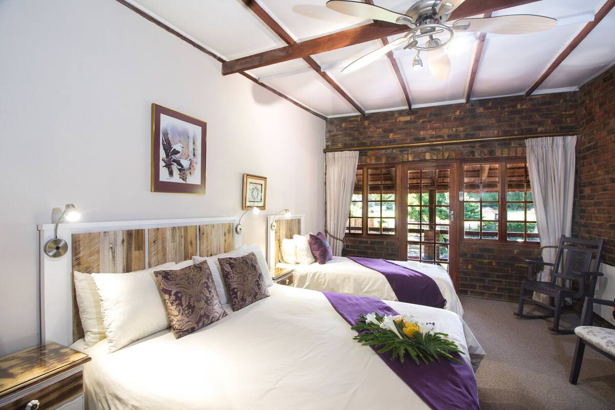 Quarto com duas camas, ventoinha no teto, janela com cortinas e parede de tijolos.