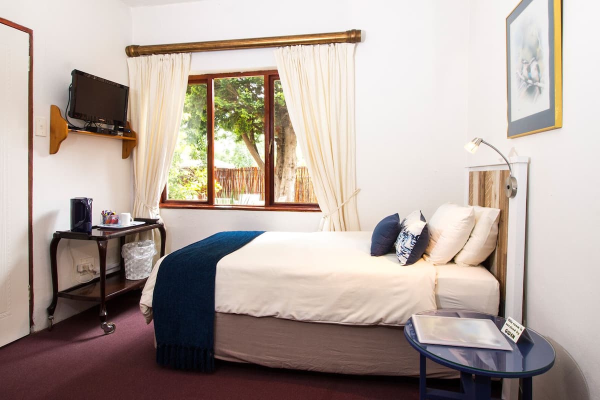 Quarto com cama, escrivaninha, cadeira, televisão e janela com cortinas.