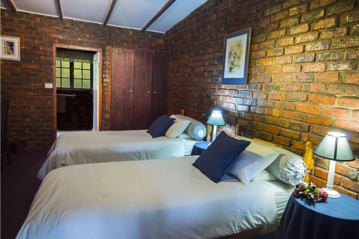 Quarto com duas camas, porta de madeira, parede de tijolos e uma pequena mesa com um abajur e flores.