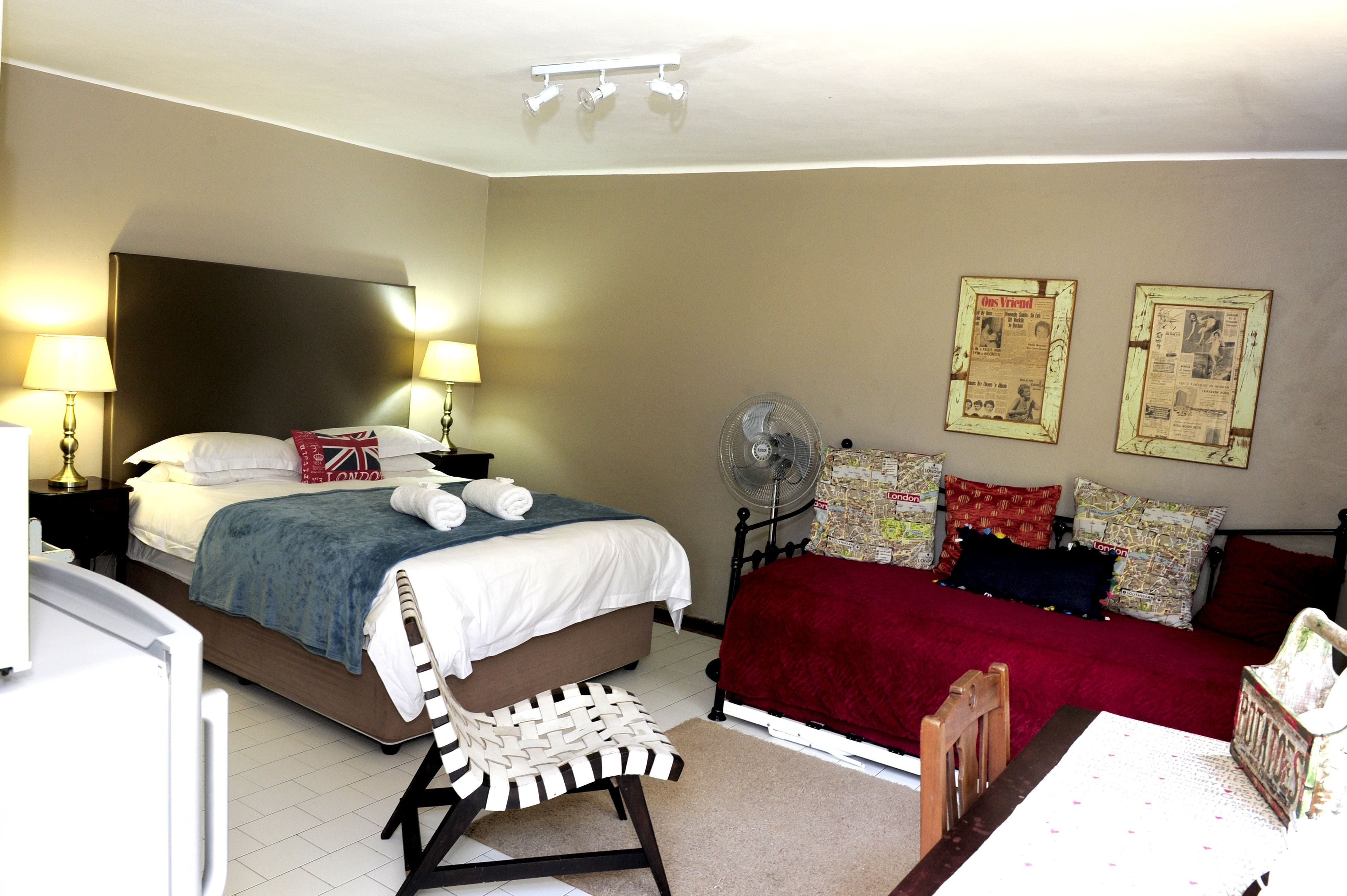 Quarto de hotel com cama, abajures, um ventilador, uma cadeira, uma mesa e dois quadros na parede.