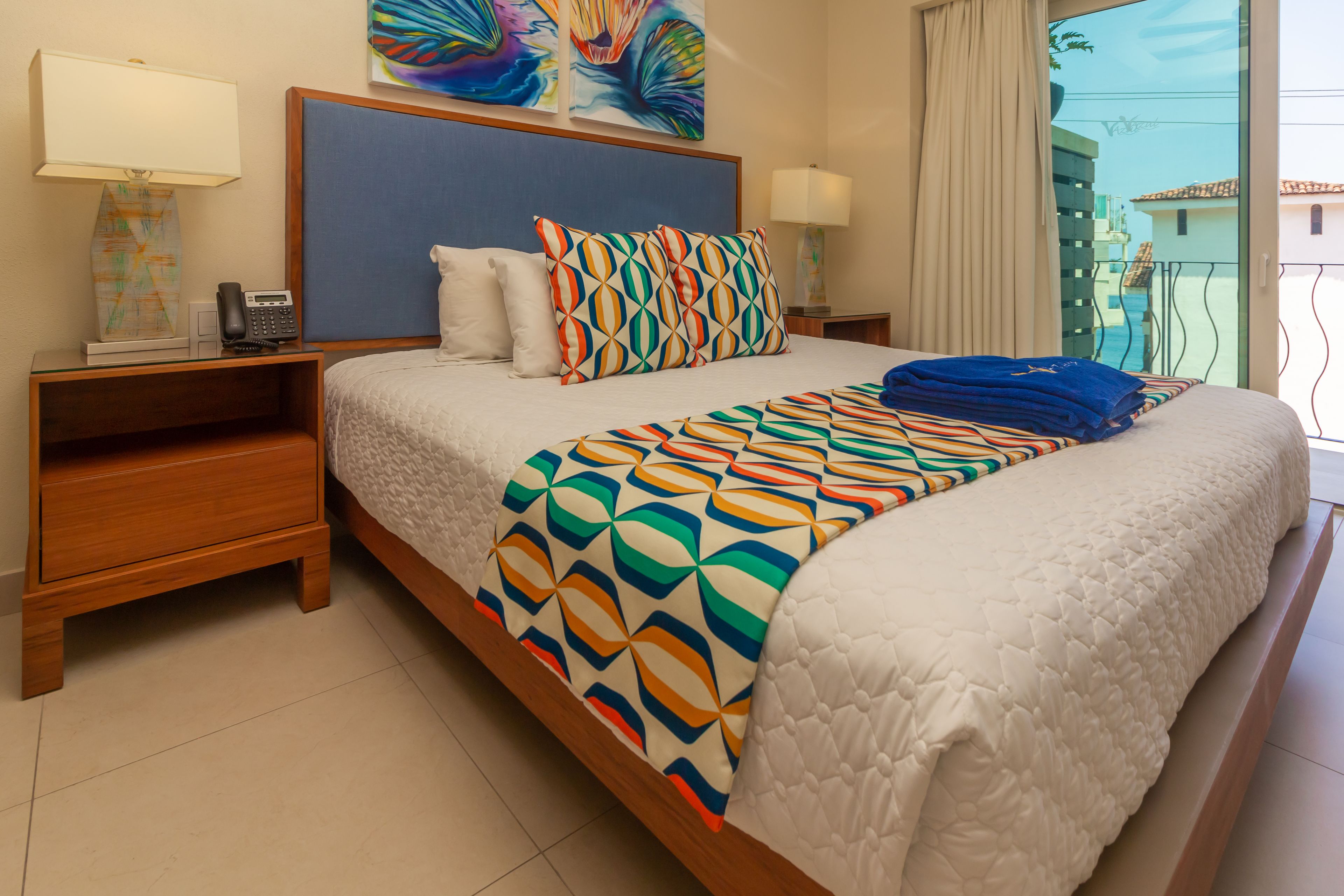 Quarto de hotel com uma cama grande, mesa de cabeceira com luminária, telefone e vista da cidade por uma varanda.