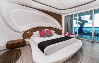 Quarto moderno com cabeceira curva, cama grande com roupa de cama branca, piso de madeira e vista para o oceano através de grandes portas de vidro deslizantes.
