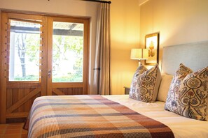 Quarto com porta de madeira, cama com almofadas estampadas, mesa de cabeceira com abajur e quadro na parede.