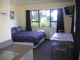 Quarto com cama, escrivaninha, cadeira, televisão e janela com cortinas.