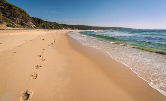 Uma praia arenosa com pegadas que conduzem à água, céu claro e paisagem acidentada ao fundo.