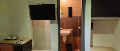 Quarto de hotel compacto com cama, escrivaninha com cadeira, TV fixada na parede, pia pequena e lixeira.