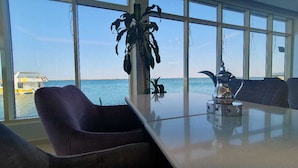 Uma sala de conferências com uma mesa grande e cadeiras, uma planta em vaso e vista para o mar através de amplas janelas.