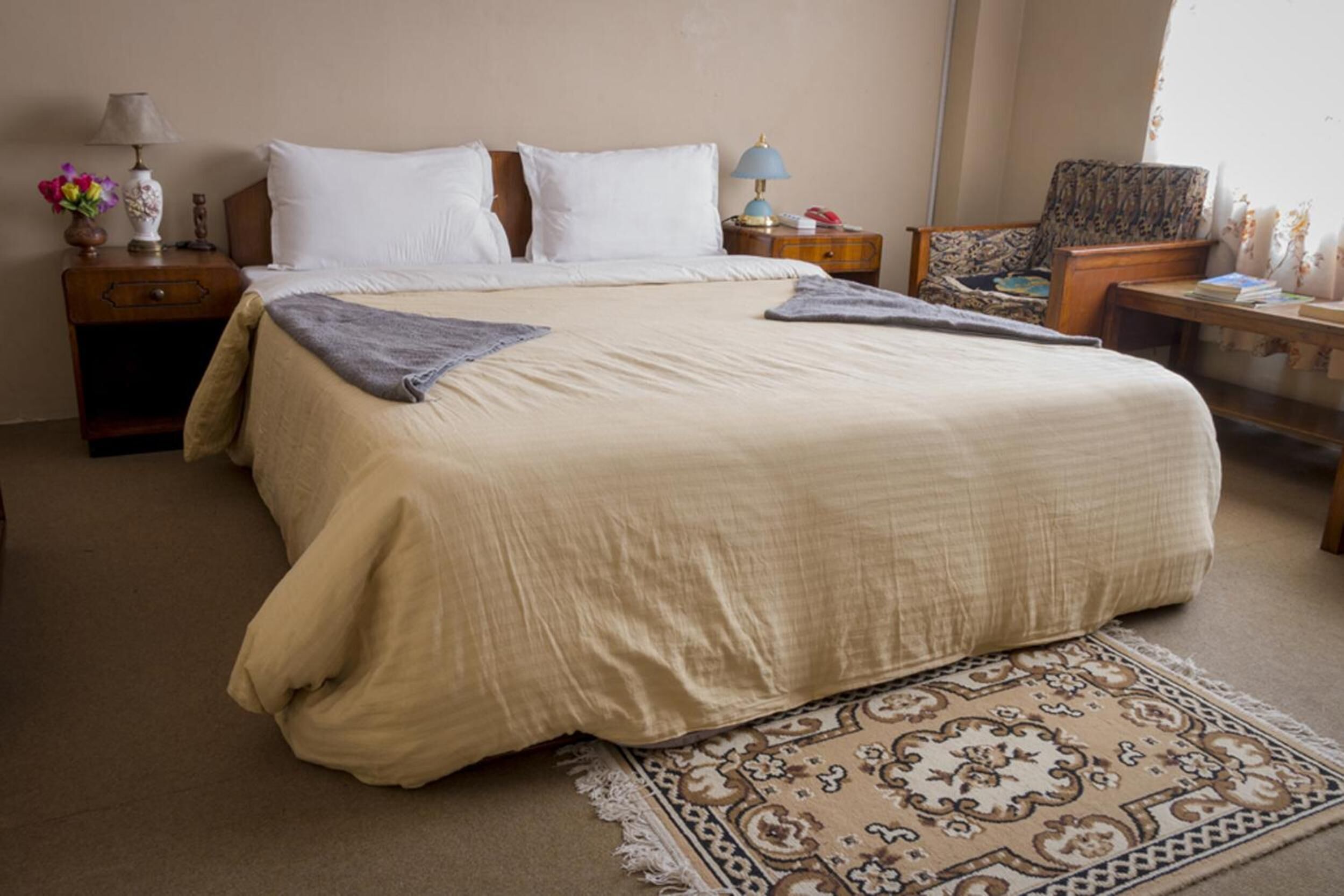 Uma cama bem arrumada com colcha e almofadas bege, uma mesa de cabeceira de madeira com abajur, um tapete estampado e uma cadeira com enchimento estampado.
