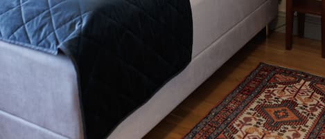 Cama bem arrumada com colcha azul, travesseiro listrado e pequena mesa de cabeceira com abajur.