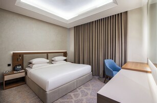 Quarto de hotel com uma cama grande, uma escrivaninha, uma cadeira, um telefone e uma janela com cortinas.