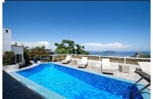 Uma piscina no terraço com água azul cristalina, cadeiras de descanso brancas e vista para o mar e paisagem distante.