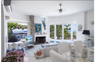 Uma sala de estar luminosa com lareira, área de jantar com mesa posta para dois e vista para o mar através de amplas janelas.
