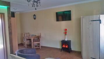 Uma sala de estar com mesa de madeira, cadeiras, sofá, televisão na parede e lareira a lenha.