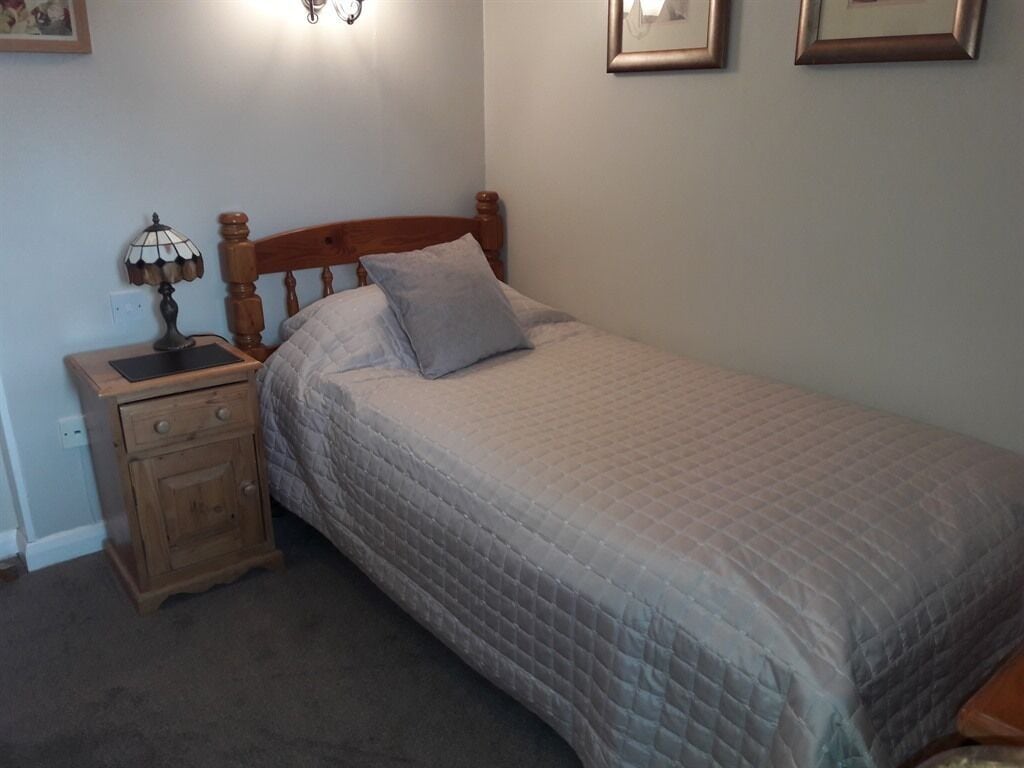 Quarto com cama de madeira, mesa de cabeceira com abajur e luminária fixada na parede.