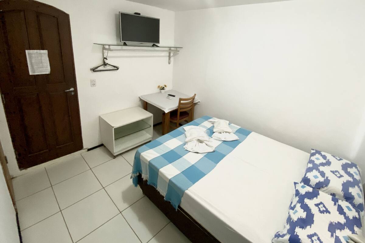 Quarto pequeno e limpo com uma cama, uma escrivaninha, uma cadeira e uma televisão em uma prateleira.