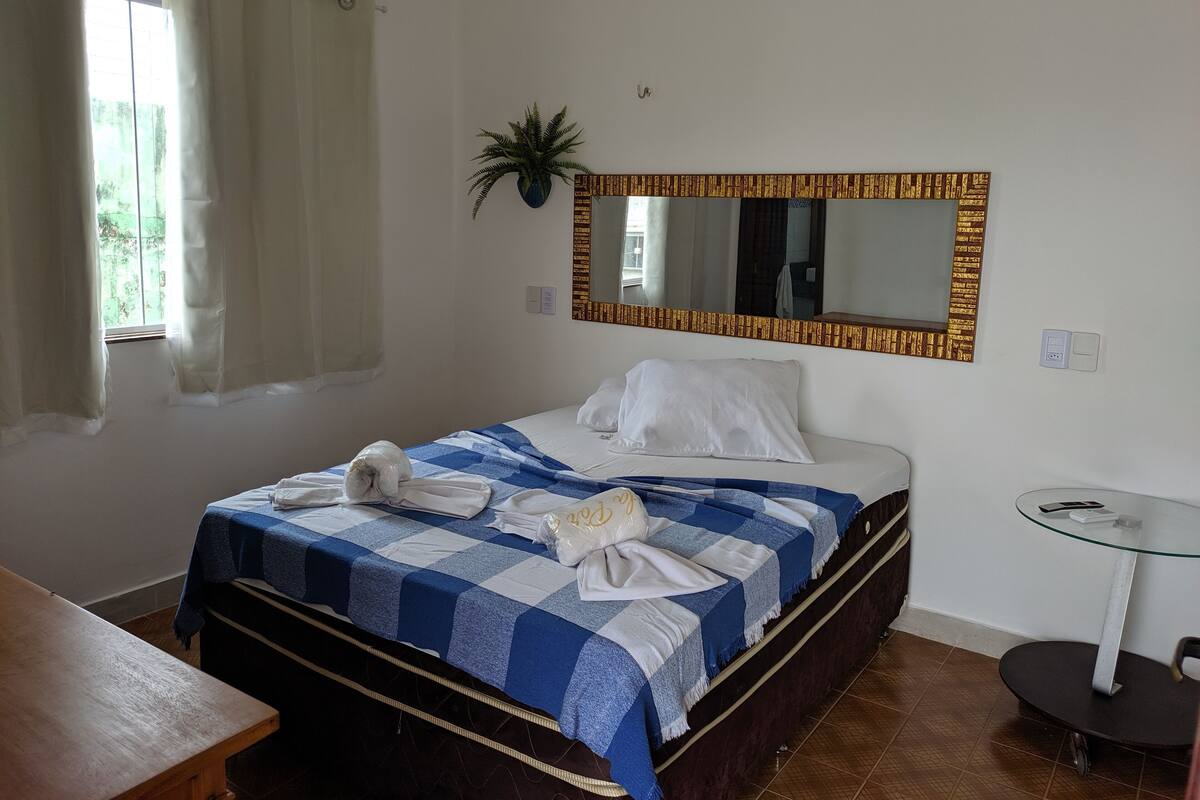 Quarto de hotel com cama, uma pequena mesa, espelho e janela com cortinas.