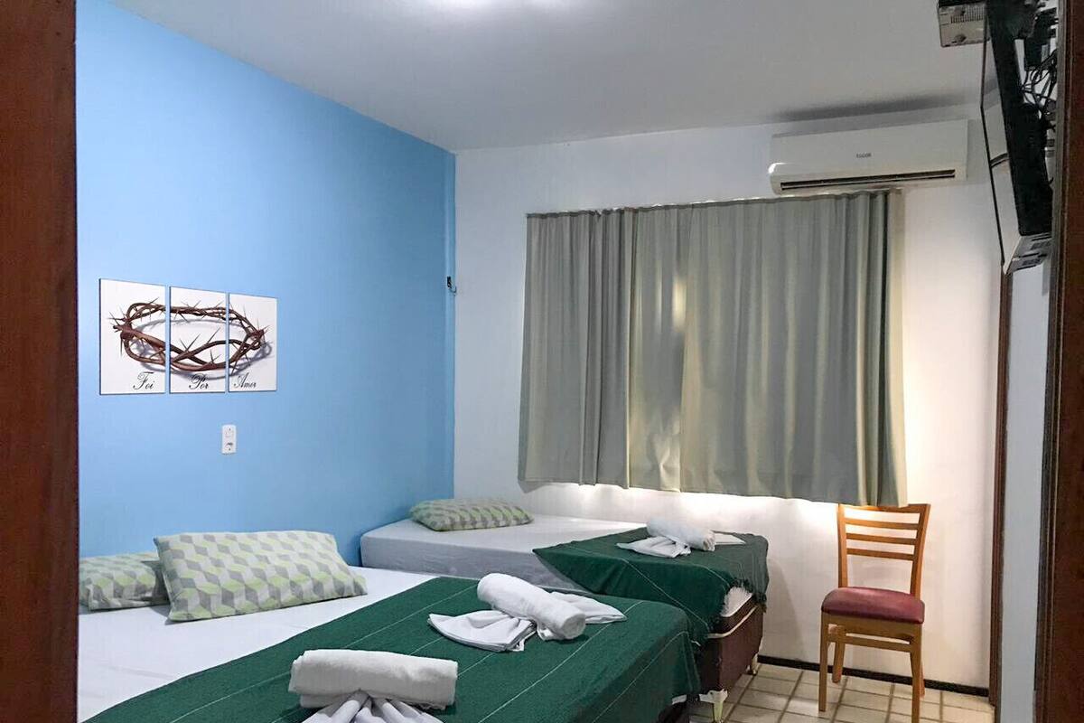 Quarto de hotel com duas camas, uma cadeira, janela com cortinas e um adorno na parede.