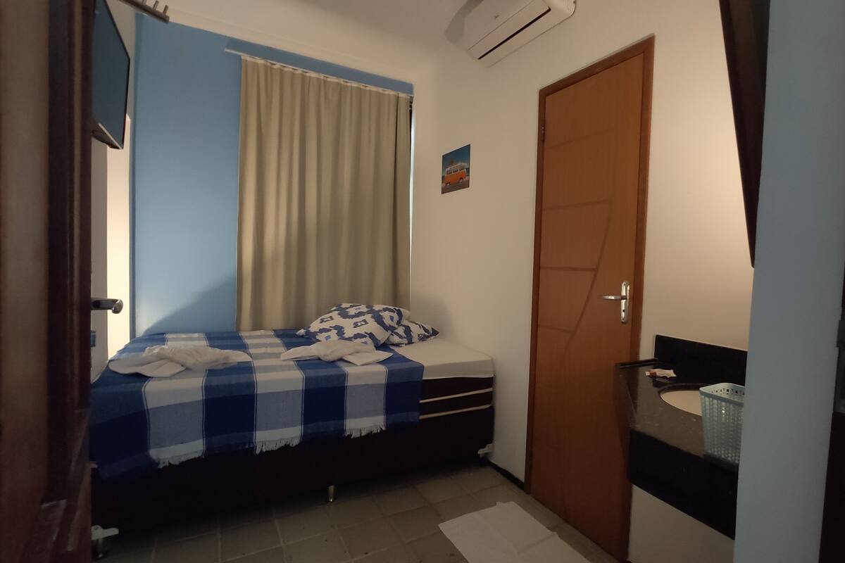Quarto pequeno e arrumado, com uma cama, uma porta e uma janela com cortinas.