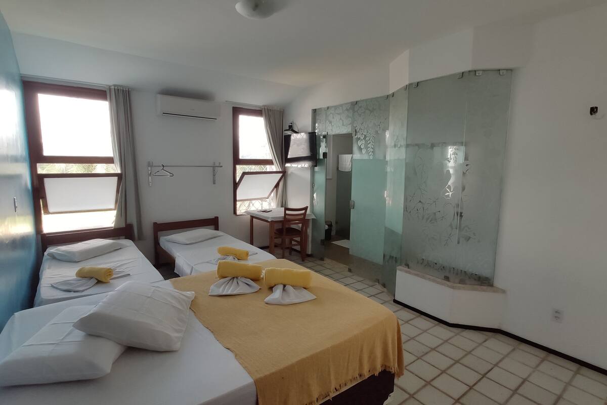 Quarto de hotel com duas camas, uma mesinha e uma cadeira. Há uma janela com cortinas, um ar condicionado e um painel de vidro decorativo.