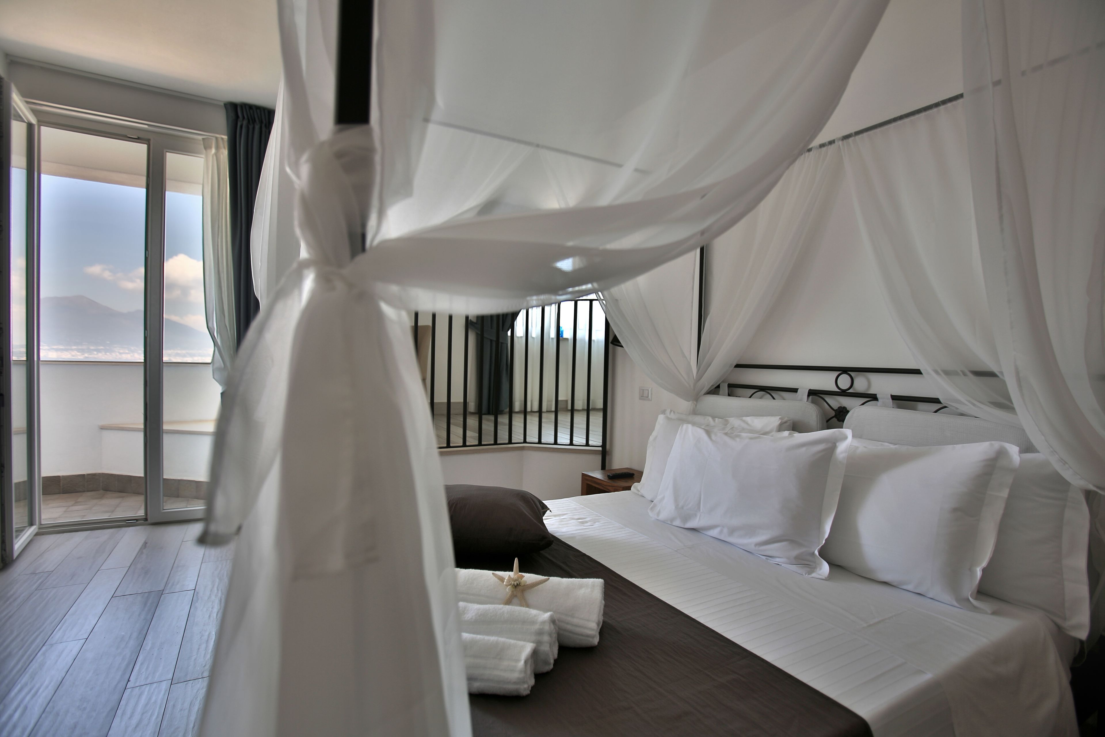 Quarto com cama, cortinas brancas e vista para o exterior.