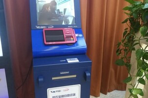 Máquina automática azul com um aparelho rosa no topo, uma placa com texto e uma planta ao lado.