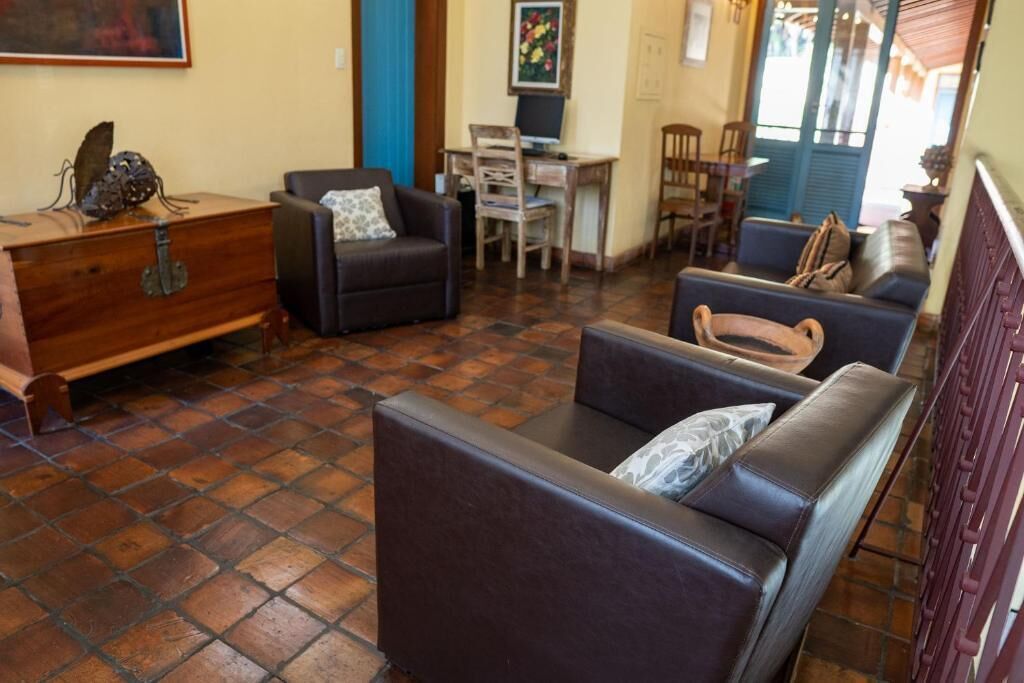 Sala de estar com sofá de couro marrom, mesa de centro de madeira e mesa lateral de madeira com uma coruja decorativa. Há uma escrivaninha de madeira com cadeira e computador.