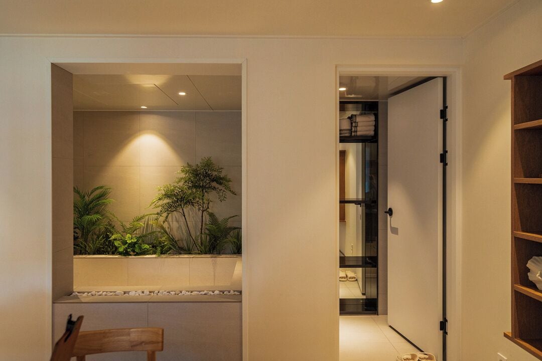Um corredor com um banco, uma porta para um quarto com uma cama e uma estante com toalhas dobradas.