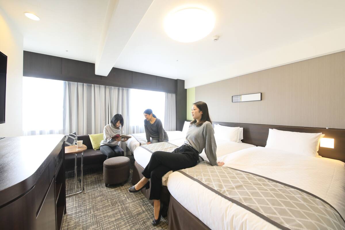 Quarto de hotel com uma cama grande, um sofá pequeno, uma escrivaninha e uma janela com cortinas.