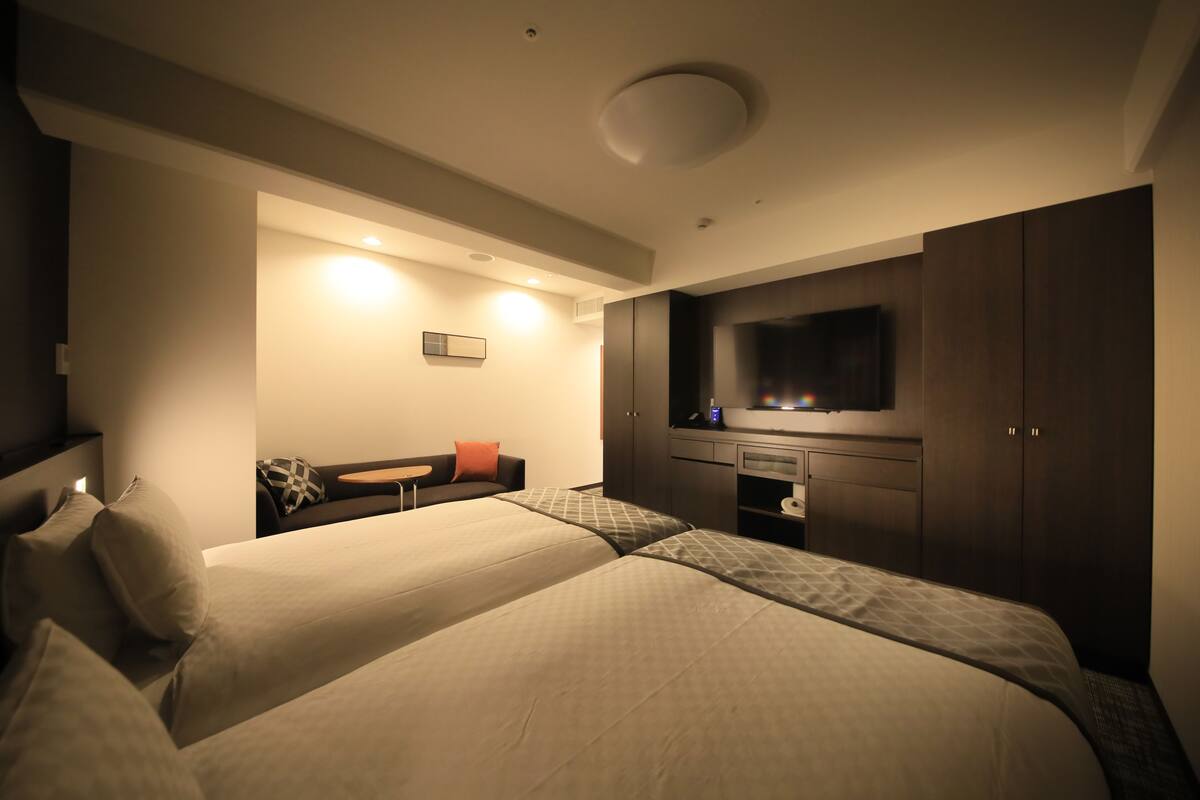 Quarto de hotel moderno com duas camas, uma televisão de tela plana, uma escrivaninha e uma área de estar com sofá e cadeiras.
