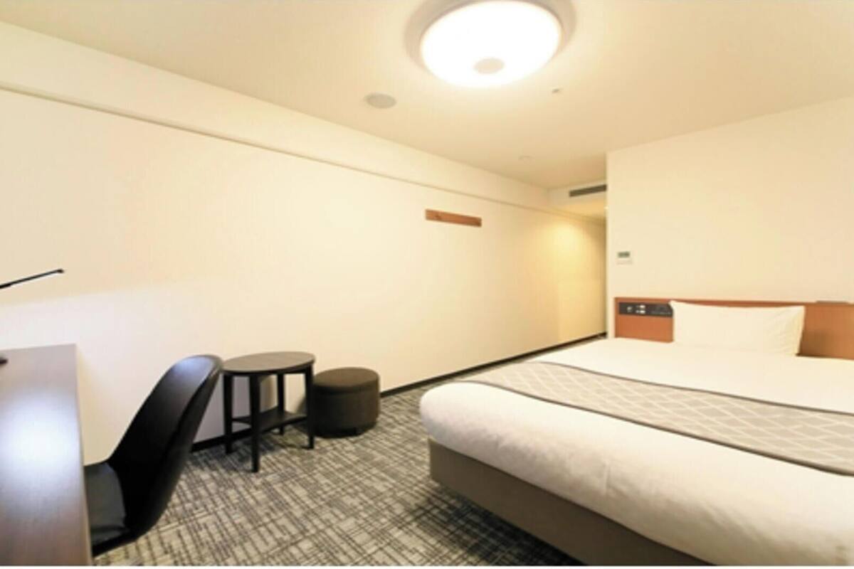 Quarto de hotel com uma cama, uma escrivaninha, uma cadeira e um banquinho.